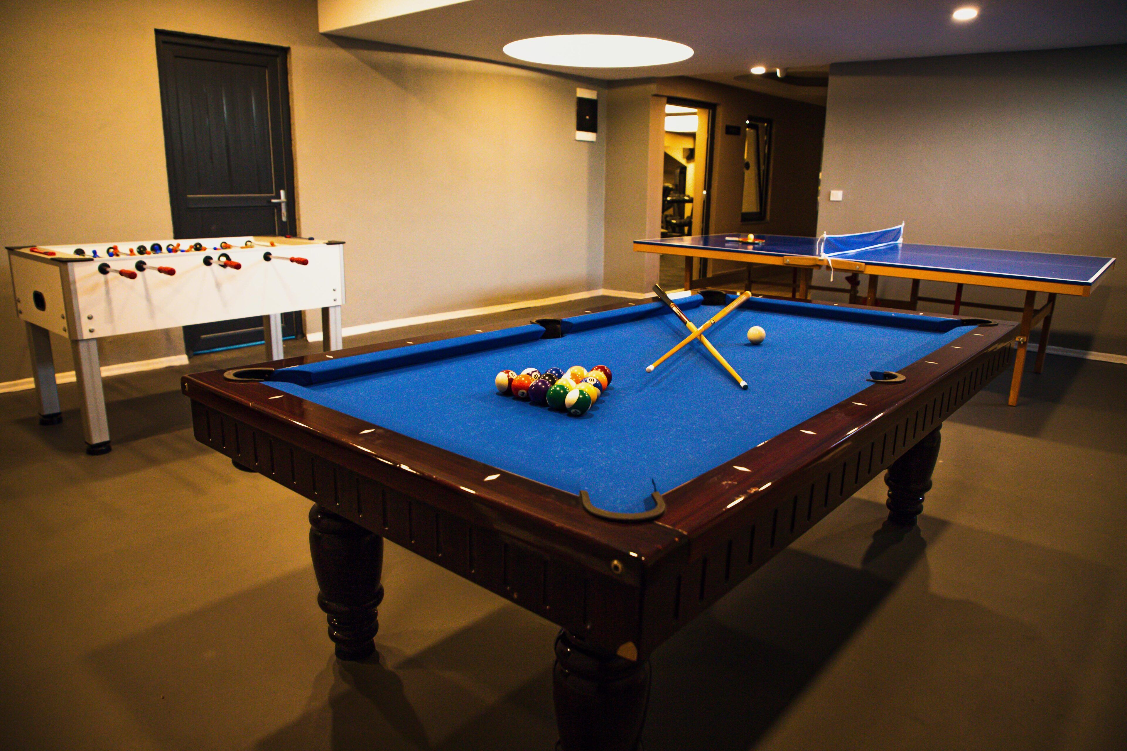Uma mesa de bilhar com bolas e tacos, uma mesa de futebol de mesa e uma mesa de pingue-pongue em um espaço com paredes bege e uma porta ao fundo.