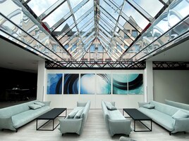 Um lounge moderno com teto de vidro, sofás cinza claro e arte abstrata nas paredes.