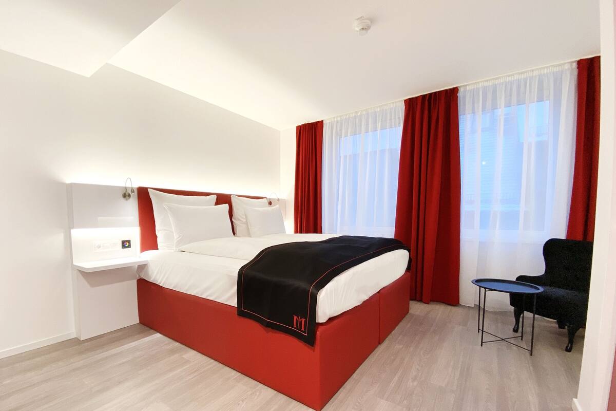 Quarto de hotel com cama, mesa de cabeceira, uma cadeira e janela com cortinas.