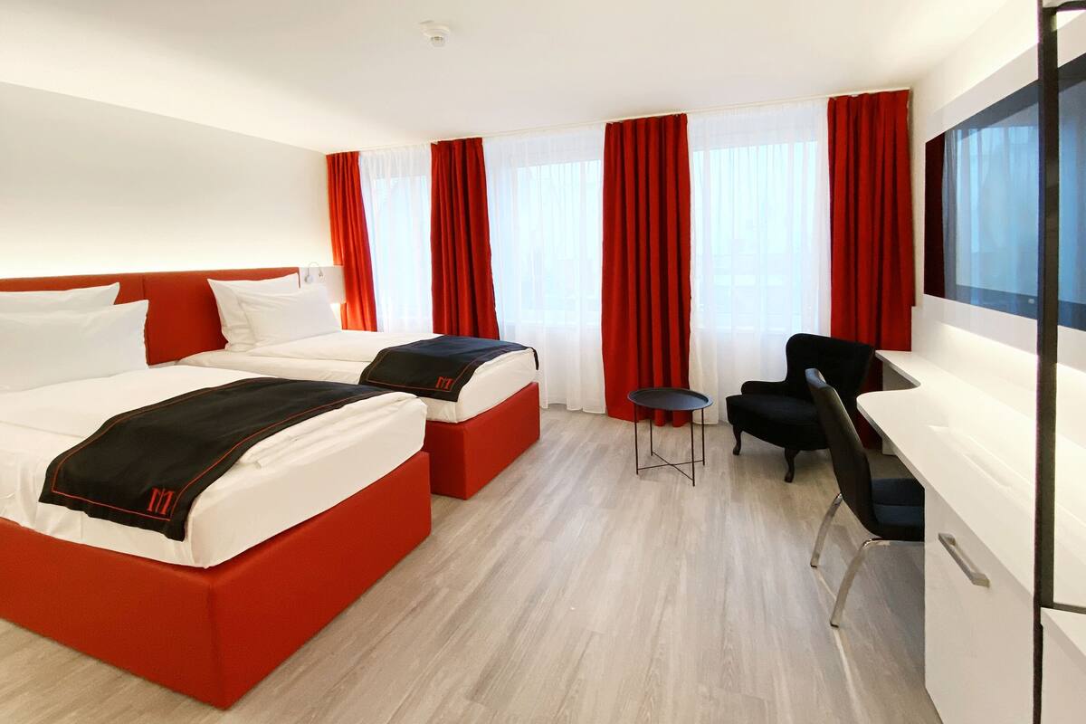 Quarto de hotel com duas camas, uma escrivaninha, uma cadeira e uma mesinha.