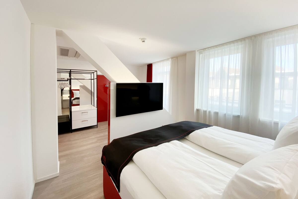 Quarto de hotel moderno com cama, televisão fixada na parede e vista para o exterior através de grandes janelas.