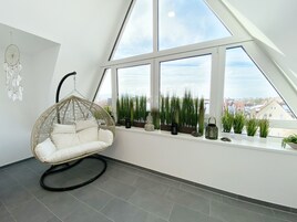 Quarto moderno com teto alto, uma cadeira suspensa, janela com vista da cidade e um conjunto de plantas em vasos.