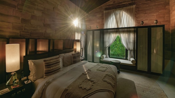Quarto com parede de pedra, cama com dossel, mesa de cabeceira, abajur e janela com cortinas.