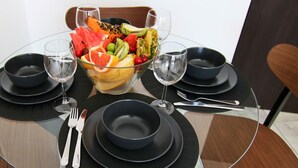 Uma mesa de jantar posta com pratos pretos, talheres e copos de vidro. Uma saladeira com frutas variadas está colocada no centro.