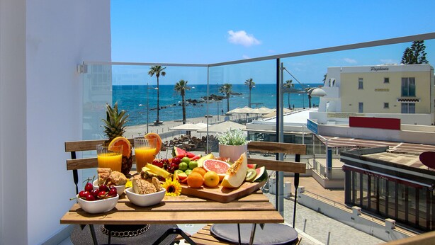 Varanda com mesa de café da manhã, incluindo frutas, pão e suco, com vista para a praia e oceano.