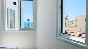 Um quarto com cama, janela com vista para o mar e edifícios, e uma porta.