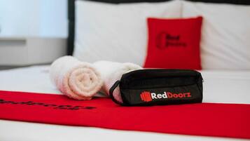 Cama bem arrumada com travesseiro vermelho e uma bolsa preta com a etiqueta 'RedDoorz' sobre um lençol branco.