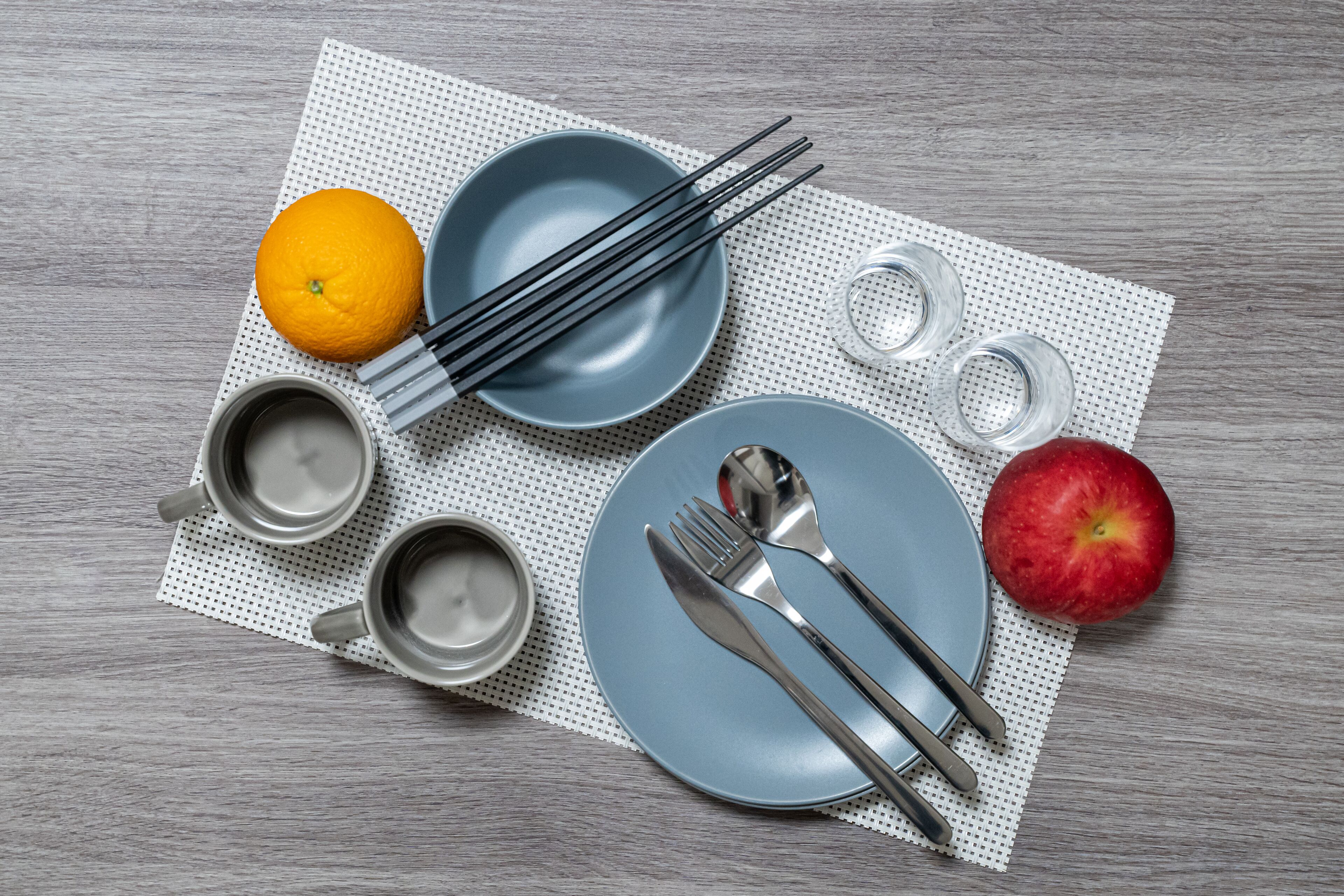 Uma mesa bem arrumada com um prato azul, talheres, hashis, uma maçã vermelha, um copo de água e uma pequena tigela de creme.