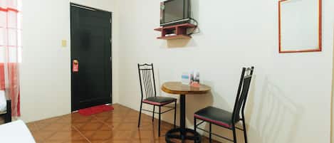 Quarto pequeno com porta preta, televisão na parede, duas cadeiras ao redor de uma mesa redonda e um tapete de entrada vermelho.