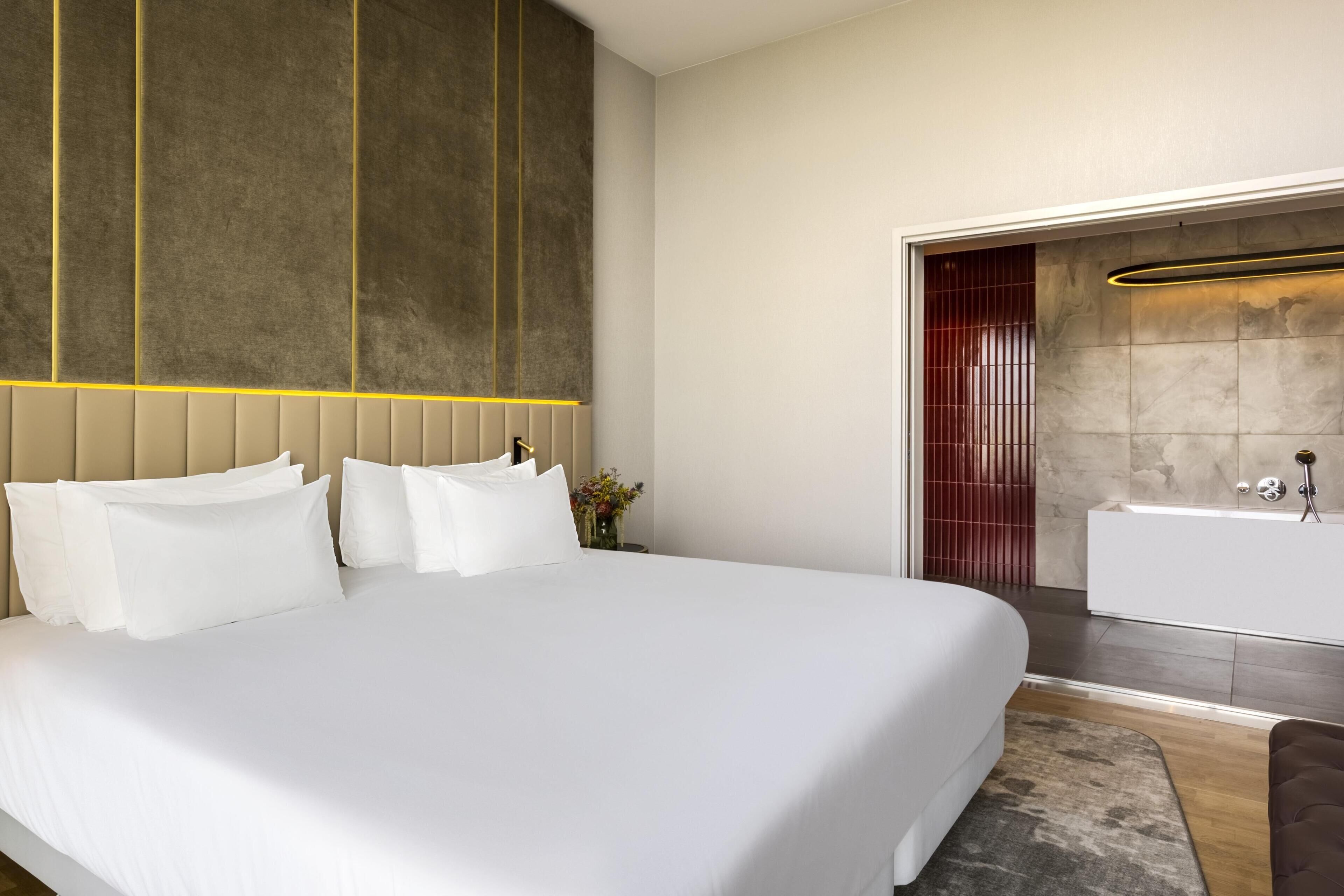 Quarto de hotel moderno com uma cama grande, um banheiro com banheira e uma porta deslizante para o banheiro.