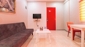 Uma sala de estar com sofá, mesa de centro com garrafas de água, TV de tela plana presa em uma parede de tijolos brancos e uma porta vermelha.