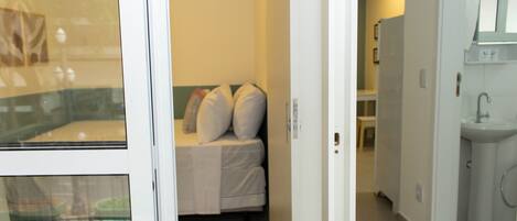 Quarto de hotel com cama, banheiro com pia e vista através de uma porta de vidro.