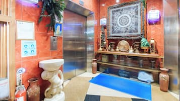 Elevador com parede de mármore, quadro emoldurado, pequena árvore de Natal e prateleira decorativa com objetos.
