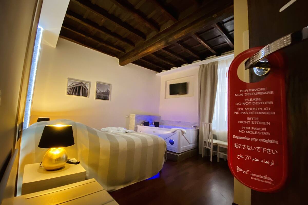 Quarto de hotel com cama, luminária de cabeceira, janela com cortinas, mesa pequena com luminária e placa vermelha com texto em vários idiomas.