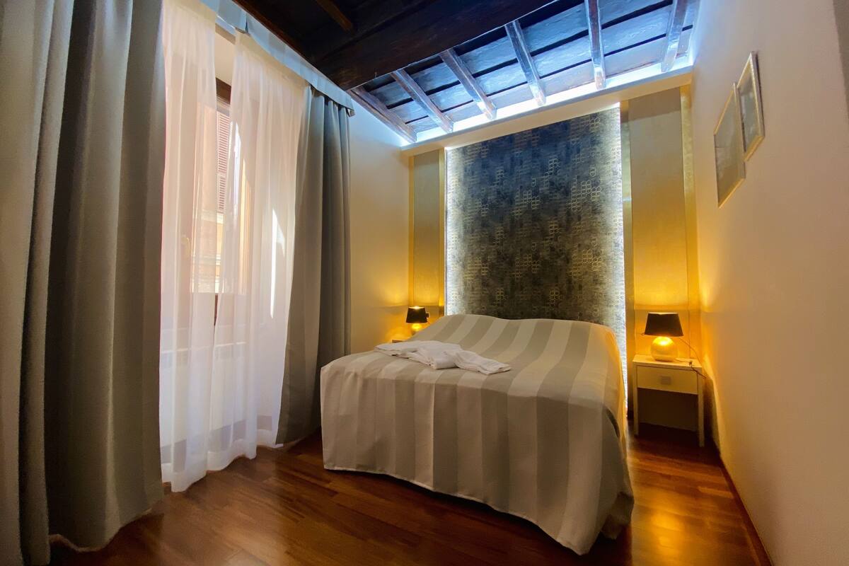 Um quarto com uma cama, duas luminárias de cabeceira, uma janela com cortinas e um claraboia.