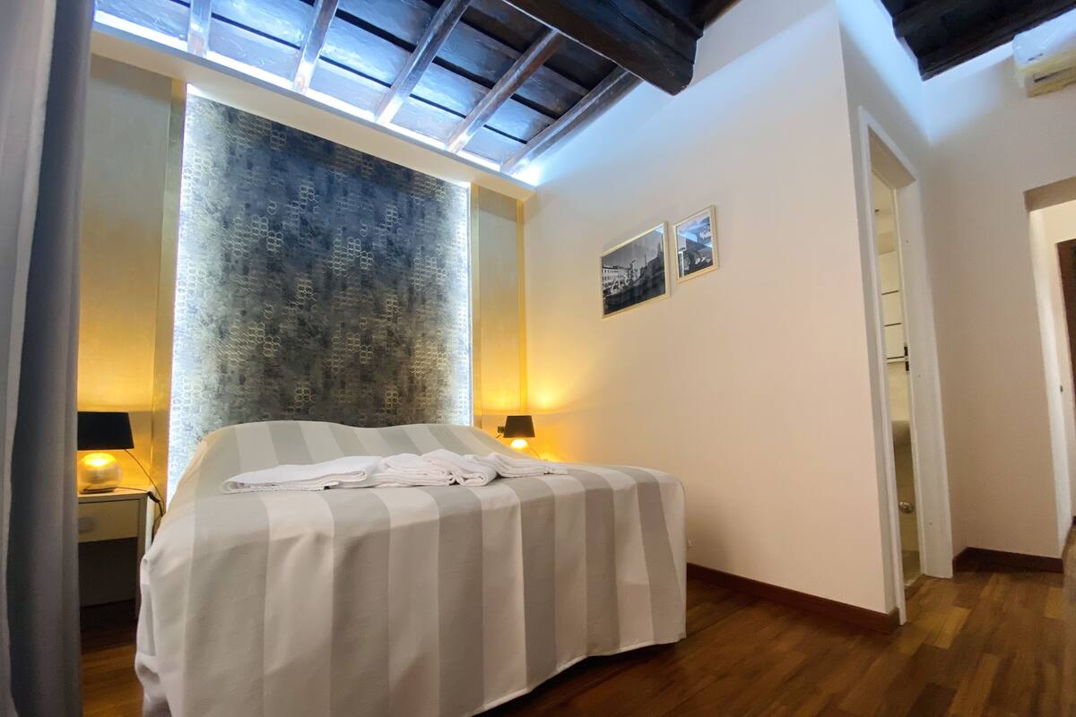 Quarto de hotel com uma cama, duas luminárias de cabeceira, uma janela grande com vista e uma porta que dá para outro espaço.
