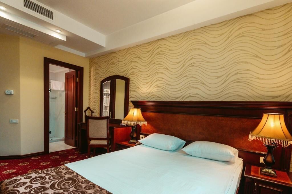 Quarto de hotel com uma cama grande, duas luminárias laterais, um espelho e um tapete estampado.