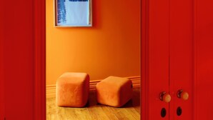Um quarto com piso de madeira, dois pufes laranja, um quadro pendurado na parede e uma porta vermelha com duas maçanetas.
