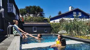 Piscina com deck de madeira e guarda-corpos de metal, cercada por vegetação e edifícios.