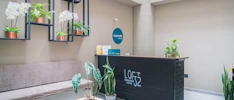 Uma área de recepção de hotel com design moderno, apresentando um balcão de atendimento, área de estar e diversos vasos de plantas.