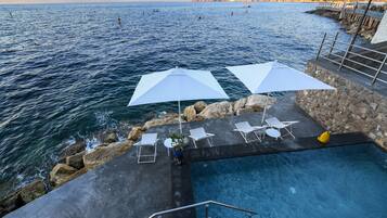 Área de piscina com guarda-sóis brancos, espreguiçadeiras e vista para o mar.