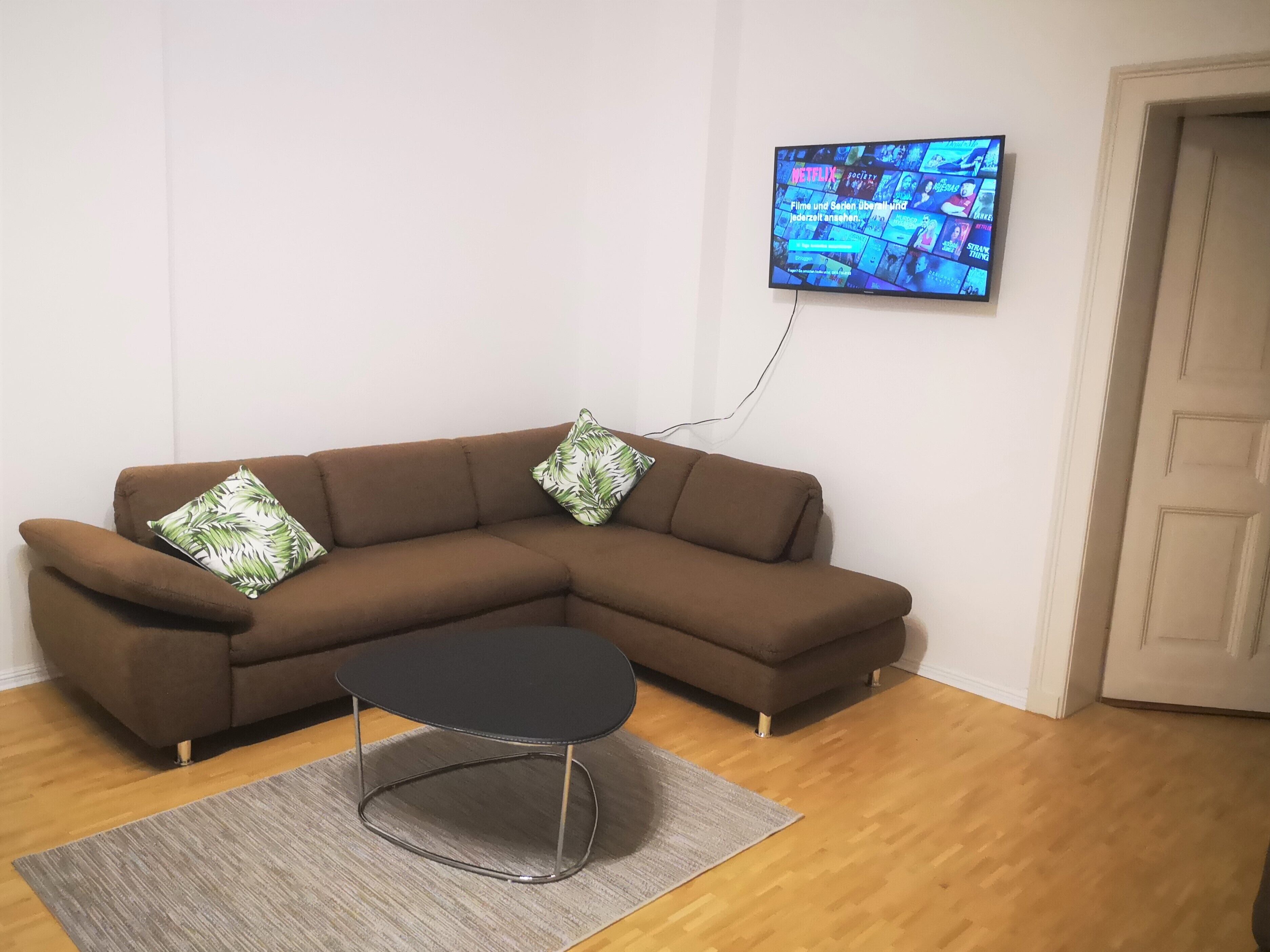 Uma sala de estar com um sofá seccional marrom, duas almofadas estampadas em verde e branco, uma mesa de centro preta com pernas de metal, um tapete cinza e uma TV de tela plana na parede exibindo a interface da Netflix.
