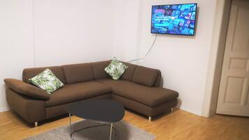 Uma sala de estar com um sofá seccional marrom, duas almofadas estampadas em verde e branco, uma mesa de centro preta com pernas de metal, um tapete cinza e uma TV de tela plana na parede exibindo a interface da Netflix.