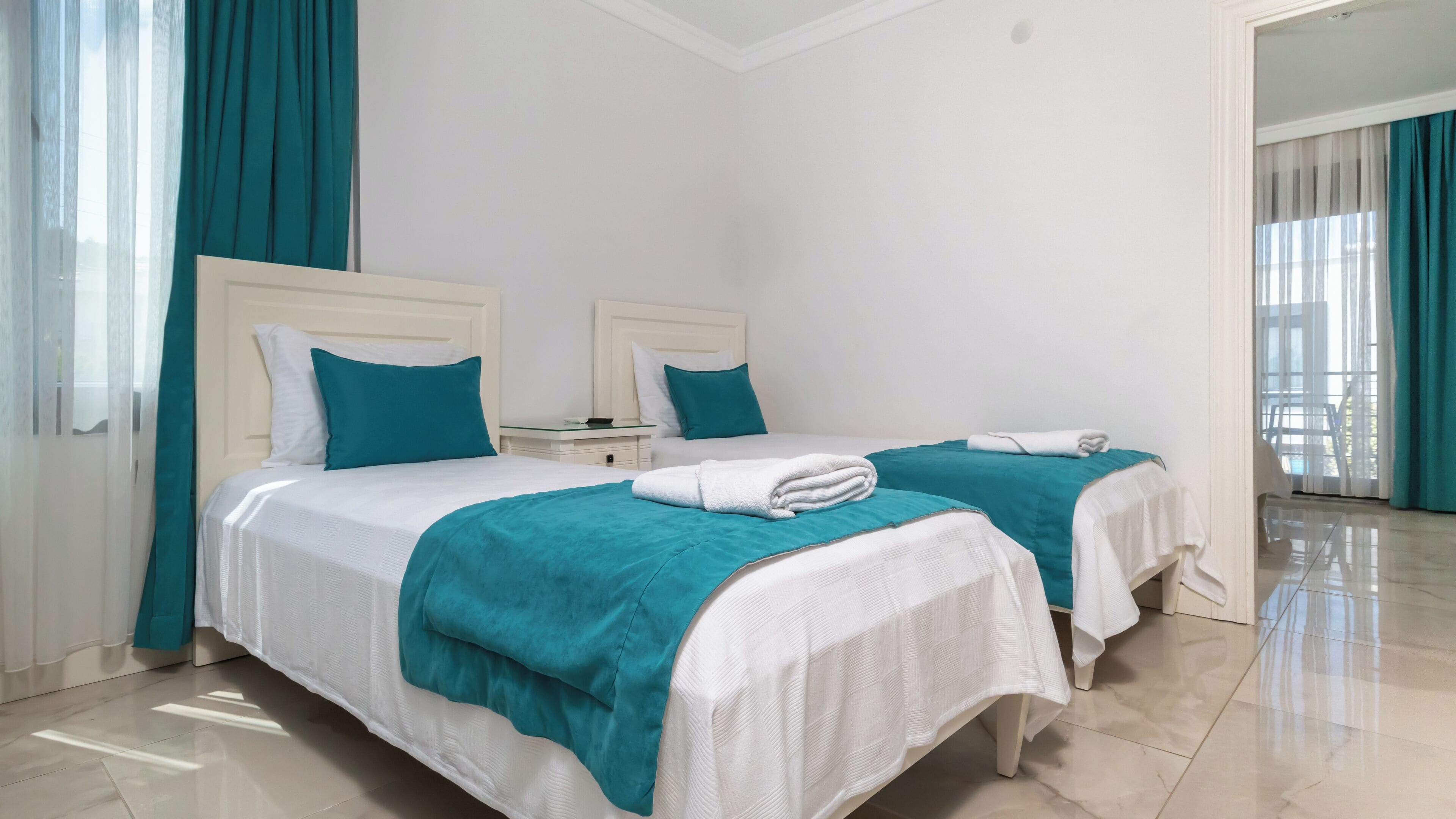 Quarto de hotel com duas camas, cada uma com roupa de cama branca e detalhes azul-turquesa, uma pequena mesa de cabeceira e vista para o exterior através de uma janela com cortinas azul-turquesa.