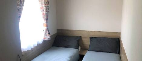 Acomodação pequena, um único quarto com duas camas individuais, uma janela com cortina e uma parede simples.
