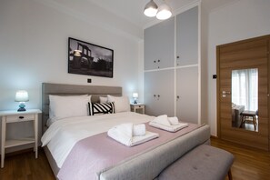 Quarto de hotel moderno com uma cama grande, duas mesas de cabeceira com luminárias, um banco cinza, um guarda-roupa de madeira e um quadro na parede.