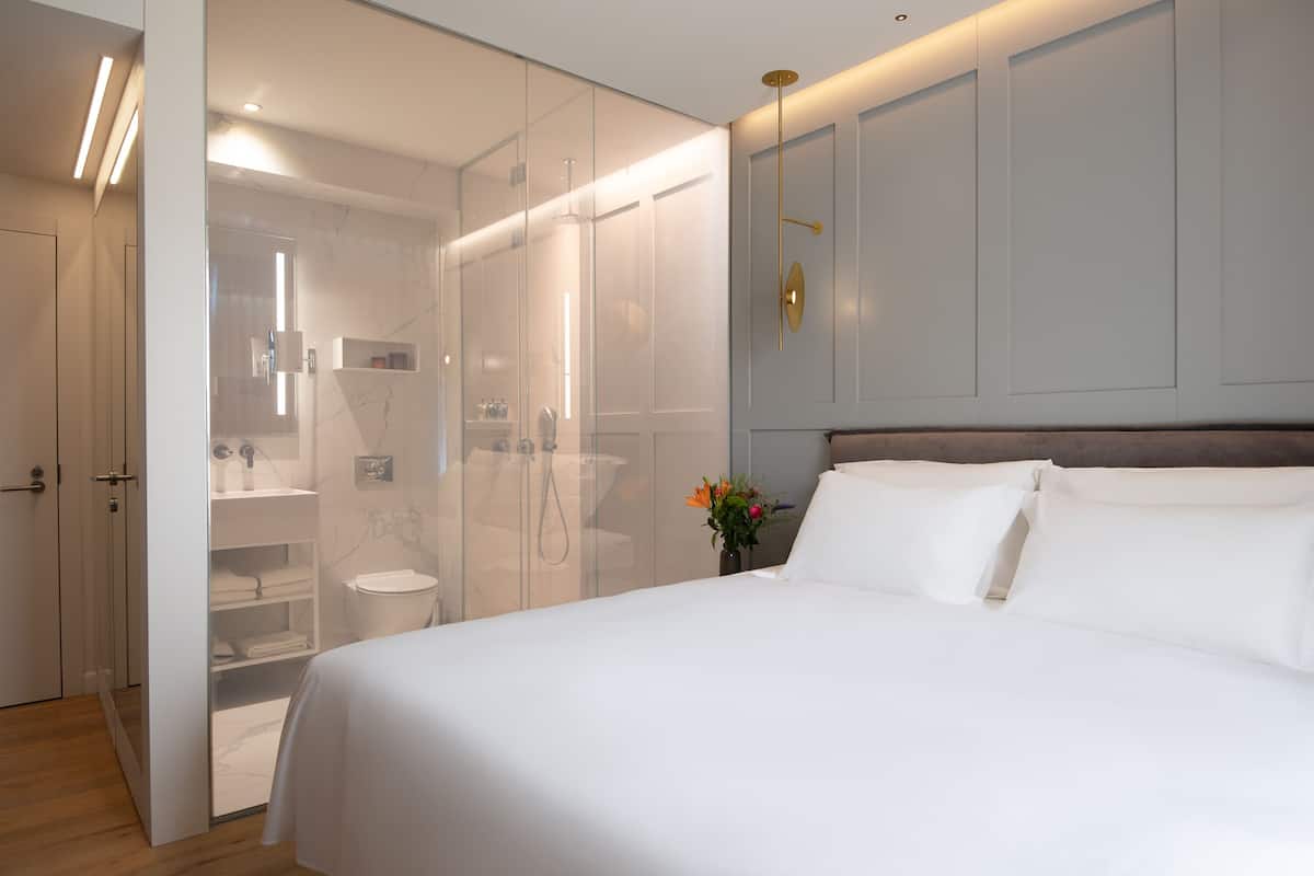 Quarto de hotel moderno com uma cama grande, um closet espaçoso, box de vidro e uma pequena mesa de cabeceira com um vaso de flores.