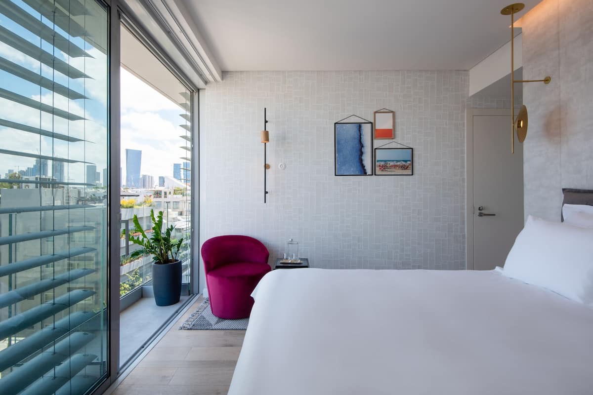 Quarto de hotel com uma cama grande, uma cadeira vermelha, uma mesinha com um recipiente de vidro e vista para a cidade através de janelas do chão ao teto.