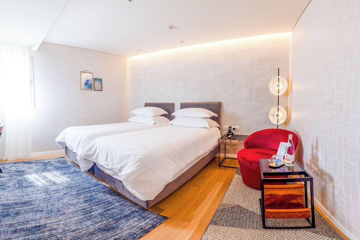 Quarto de hotel moderno com uma cama grande, uma cadeira vermelha, uma mesa pequena com uma garrafa e uma luminária na parede.