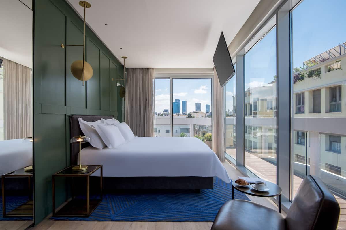 Quarto de hotel moderno com uma cama grande, uma mesinha de cabeceira, uma cadeira e vista para a cidade através de grandes janelas.