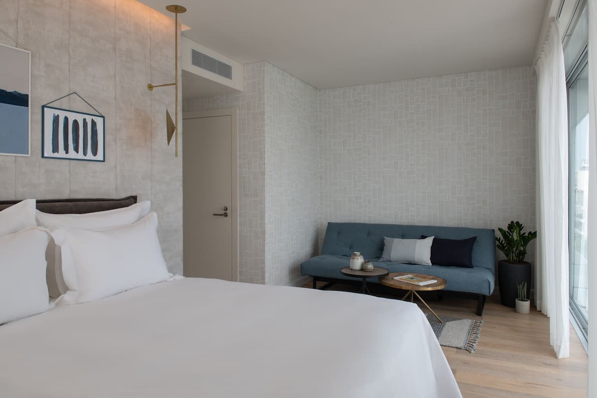 Quarto de hotel moderno com uma cama grande, um sofá, uma mesinha com um vaso e uma planta em vaso.