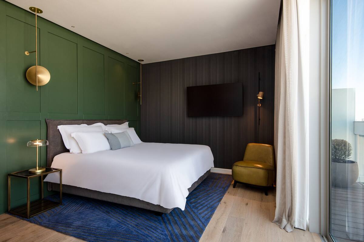 Quarto de hotel moderno com uma cama grande, uma TV de tela plana na parede, uma poltrona e vista para o exterior através de uma janela ampla.