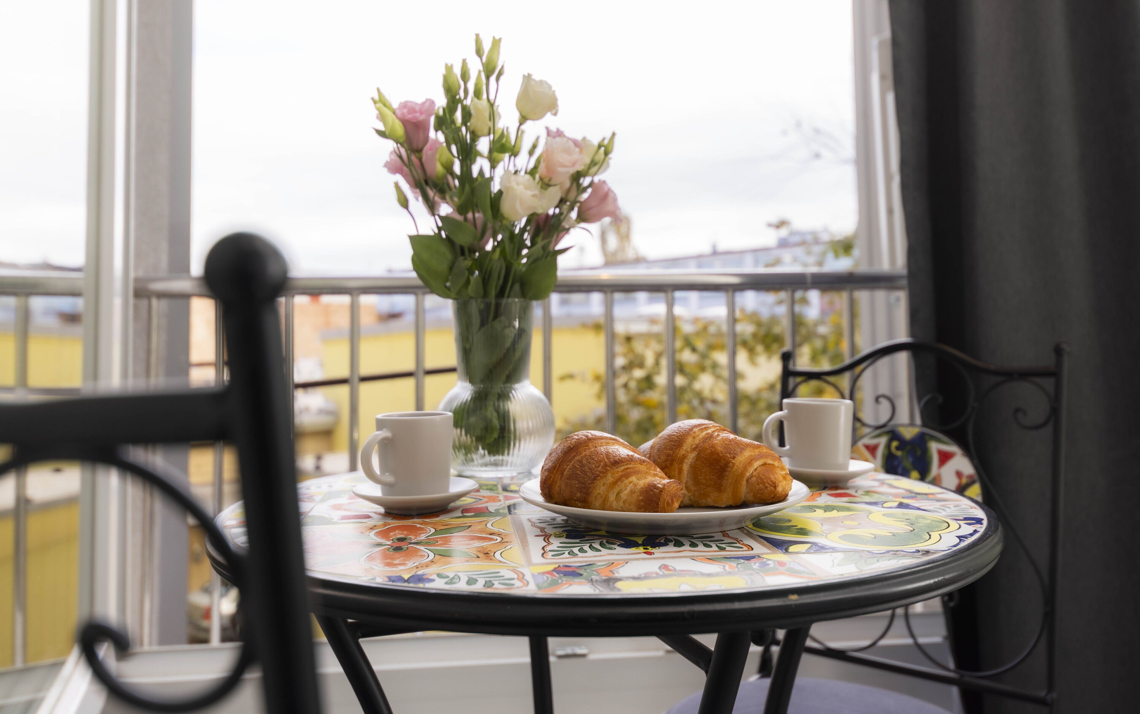 Uma mesa com dois croissants, um vaso de flores e duas chávenas numa varanda com vista para a paisagem urbana.