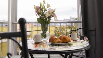 Uma mesa com dois croissants, um vaso de flores e duas chávenas numa varanda com vista para a paisagem urbana.