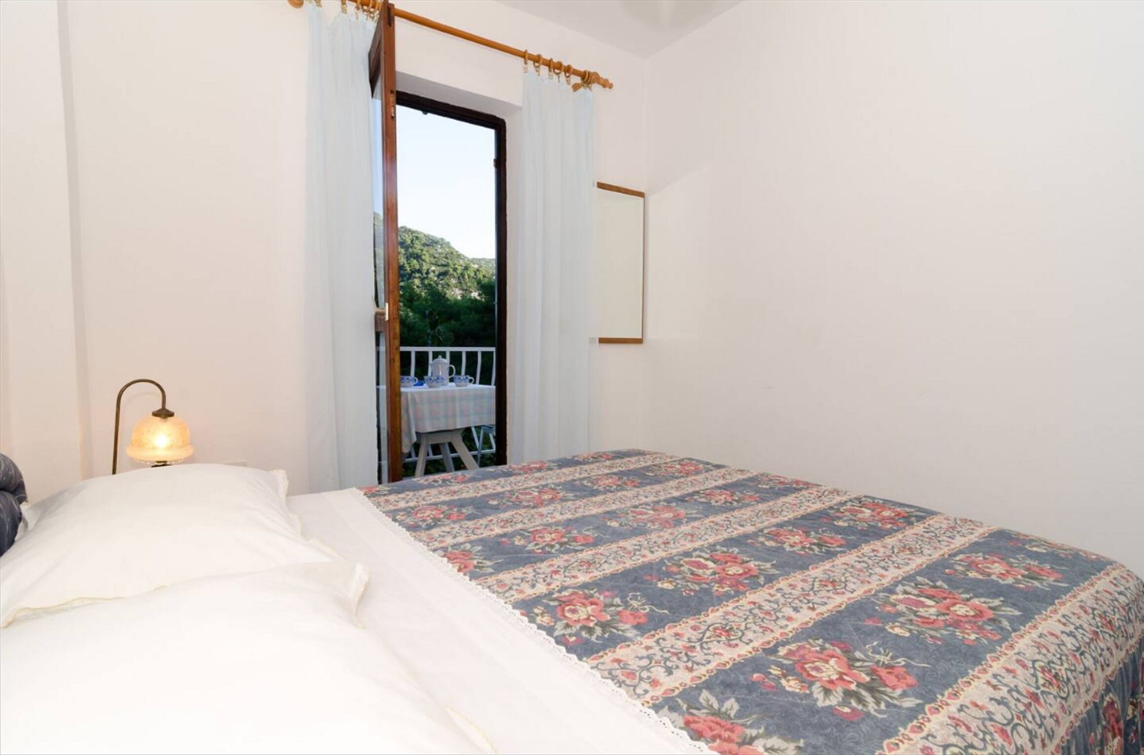 Um quarto com cama, mesa de cabeceira, luminária, janela com cortinas e vista para uma varanda com mesa e cadeiras.