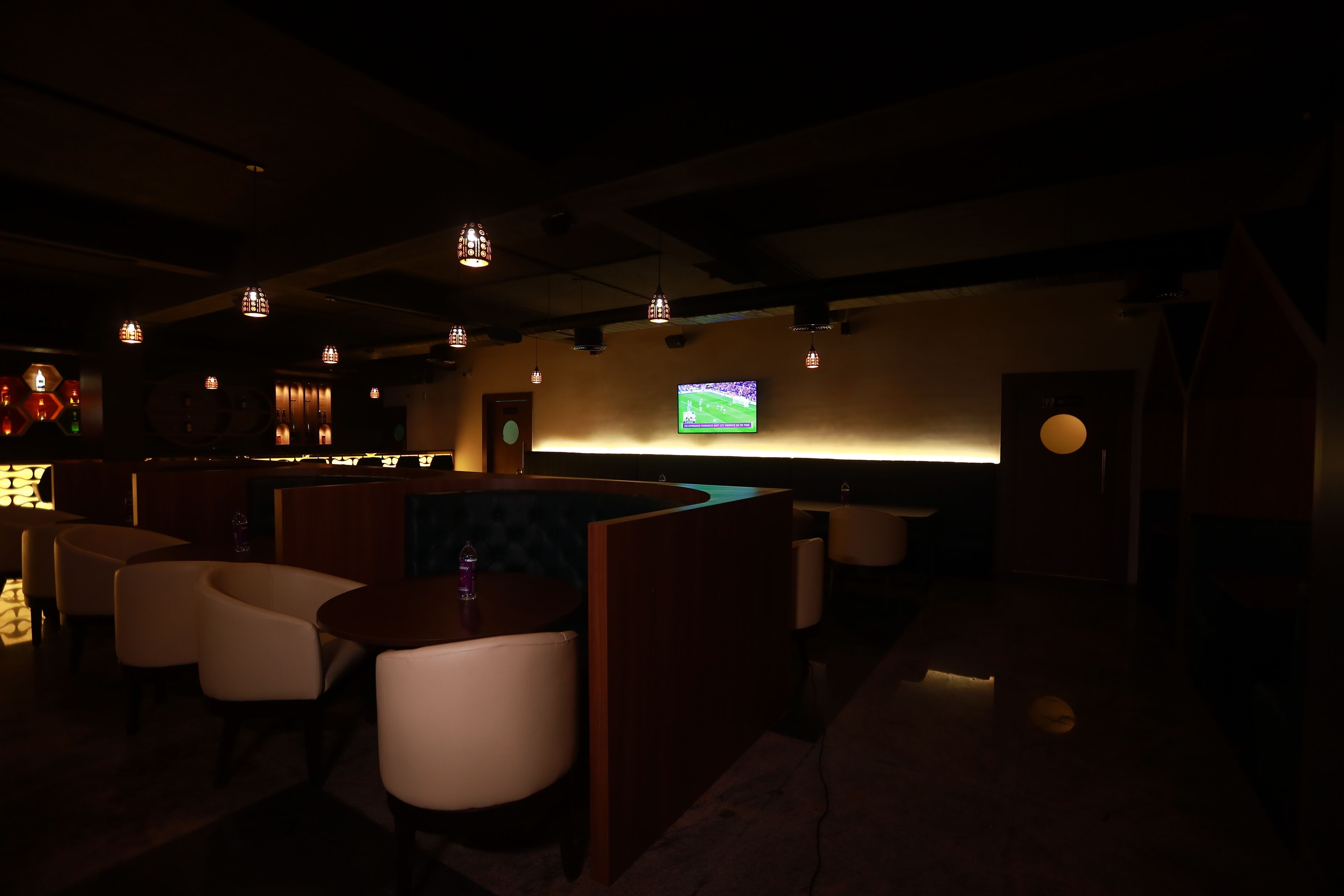 Um bar com iluminação suave, assentos e uma televisão exibindo uma partida esportiva.