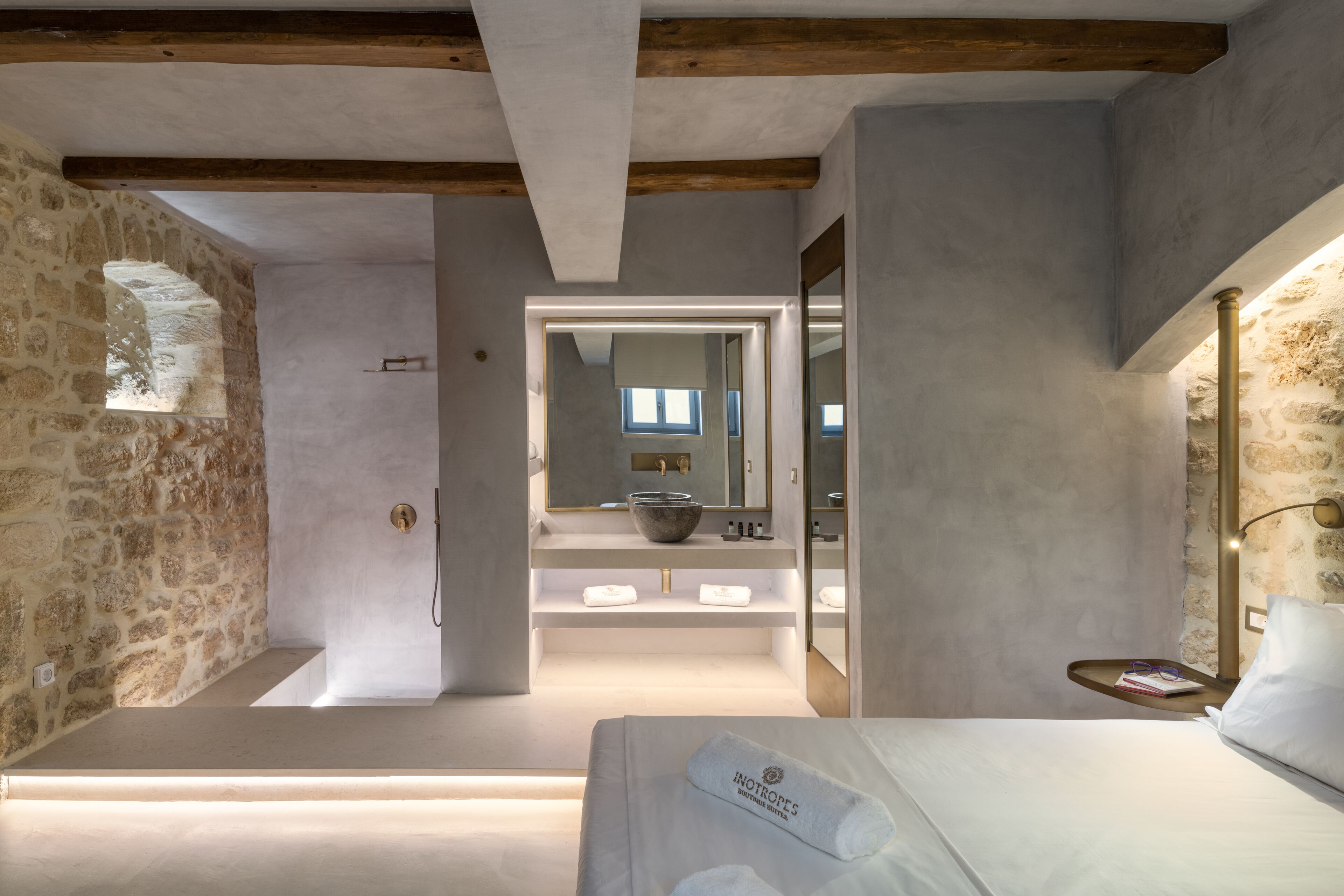 Quarto moderno com parede de pedra, uma cama grande, mesa de cabeceira com abajur e vista para um banheiro com pia de pedra.