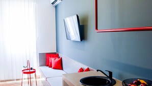 Quarto moderno com sofá, uma mesa pequena, TV na parede e área de cozinha com pia e fruteira.