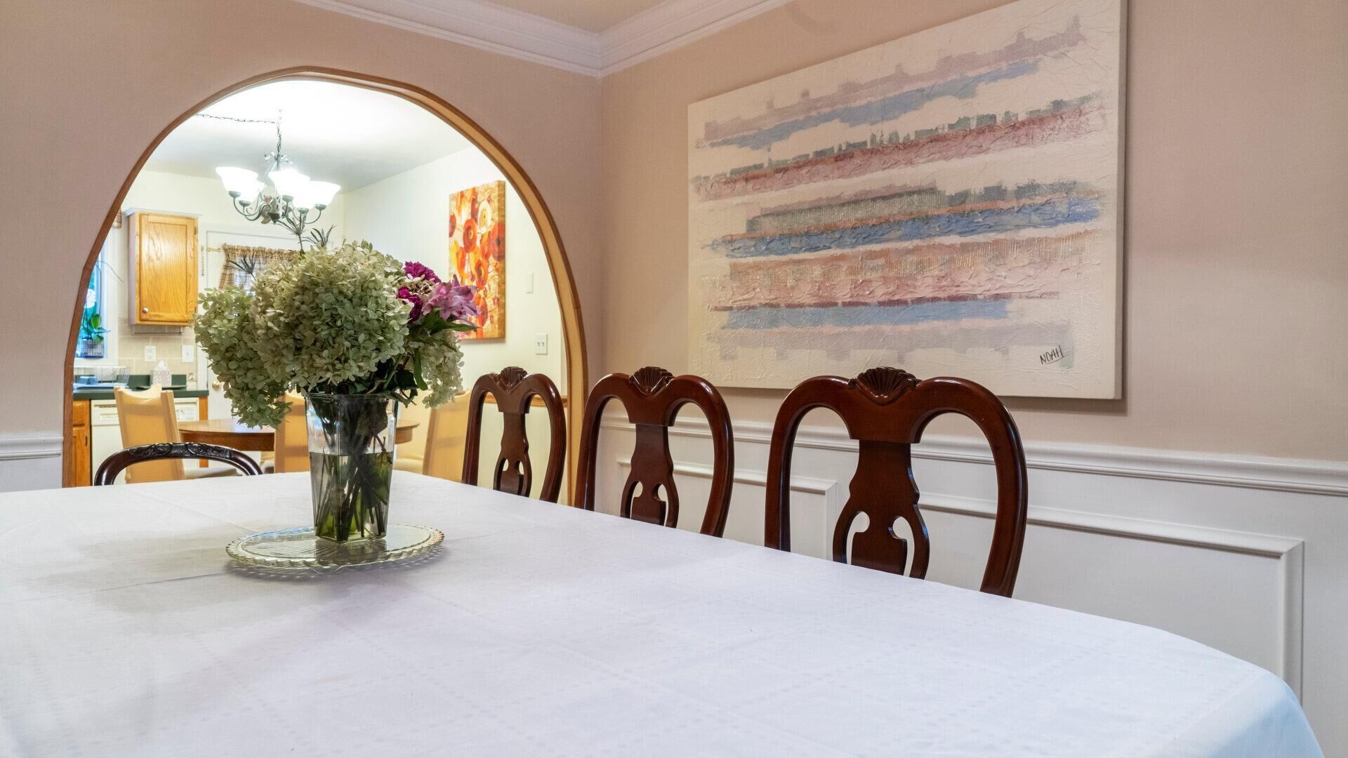 Uma sala de jantar com uma mesa posta para quatro pessoas, rodeada por cadeiras de madeira. Há um vaso com flores sobre a mesa.