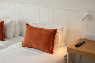Quarto de hotel com roupa de cama branca, mesa de cabeceira de madeira, controle remoto e luminária fixada na parede.