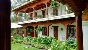 Um edifício de dois andares com varanda, rodeado por vegetação exuberante e um jardim bem cuidado.
