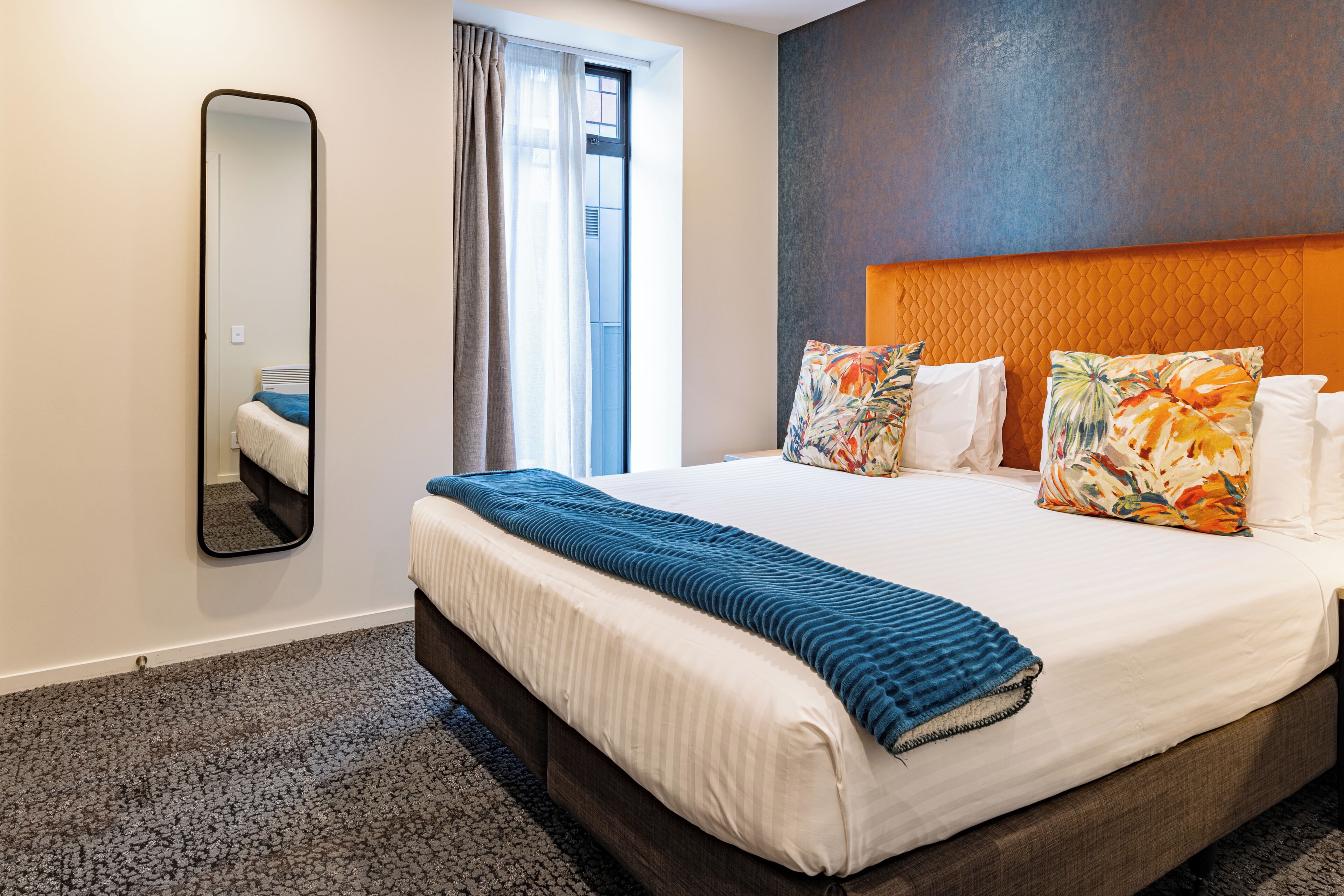Quarto de hotel com uma cama grande, um espelho, uma janela com cortinas e uma parede com textura.