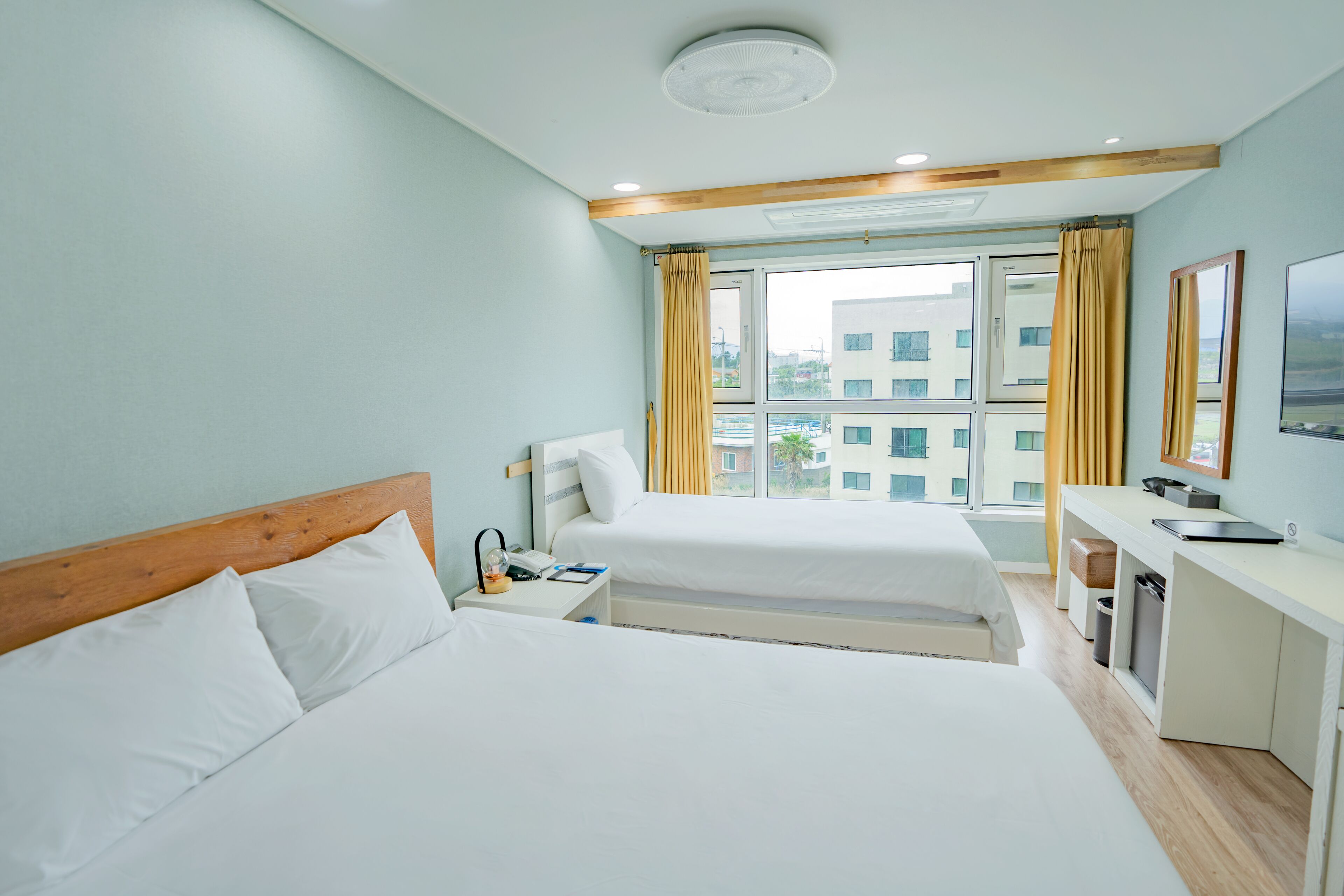 Quarto de hotel com duas camas, uma escrivaninha, uma cadeira, janela com cortinas e luminária no teto.