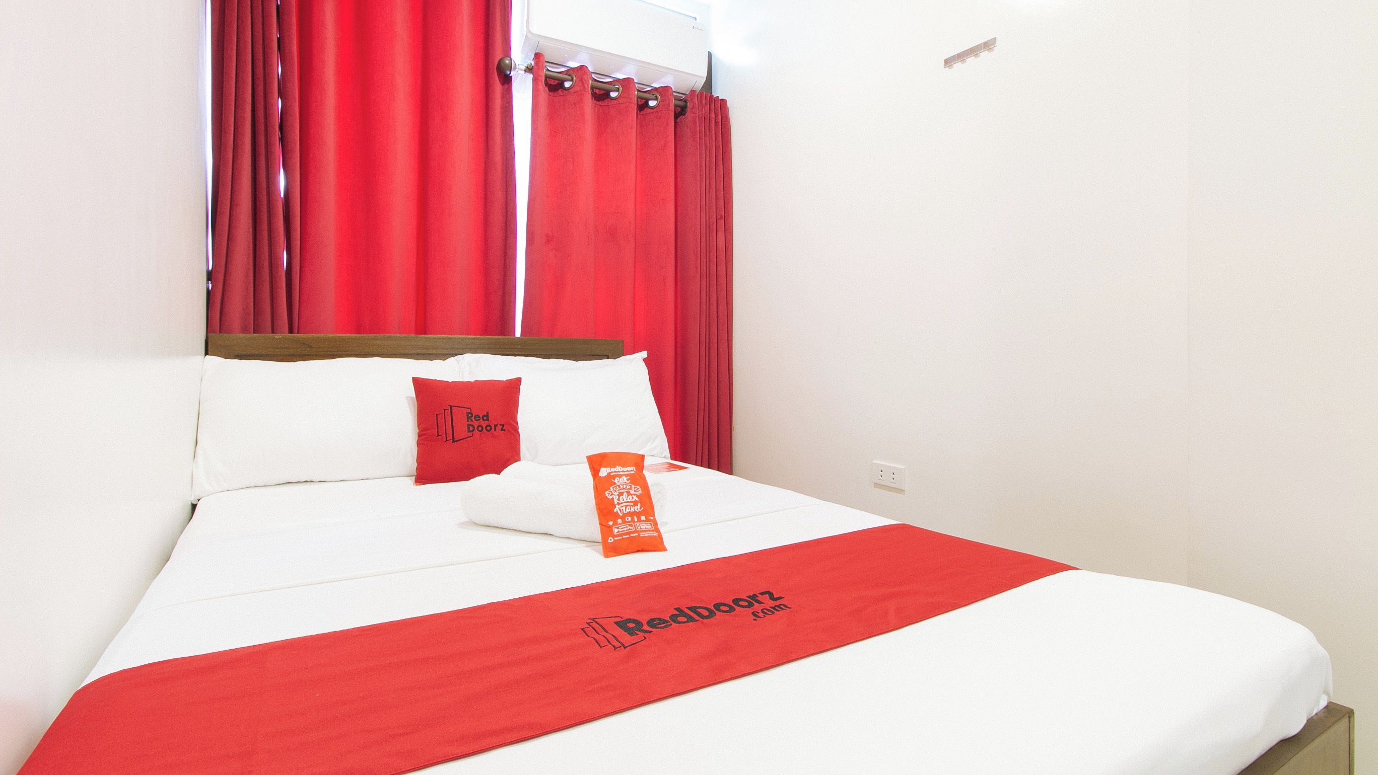 Quarto de hotel com cama, roupa de cama branca, cortinas vermelhas e edredom vermelho com o logo da RedDoorz.