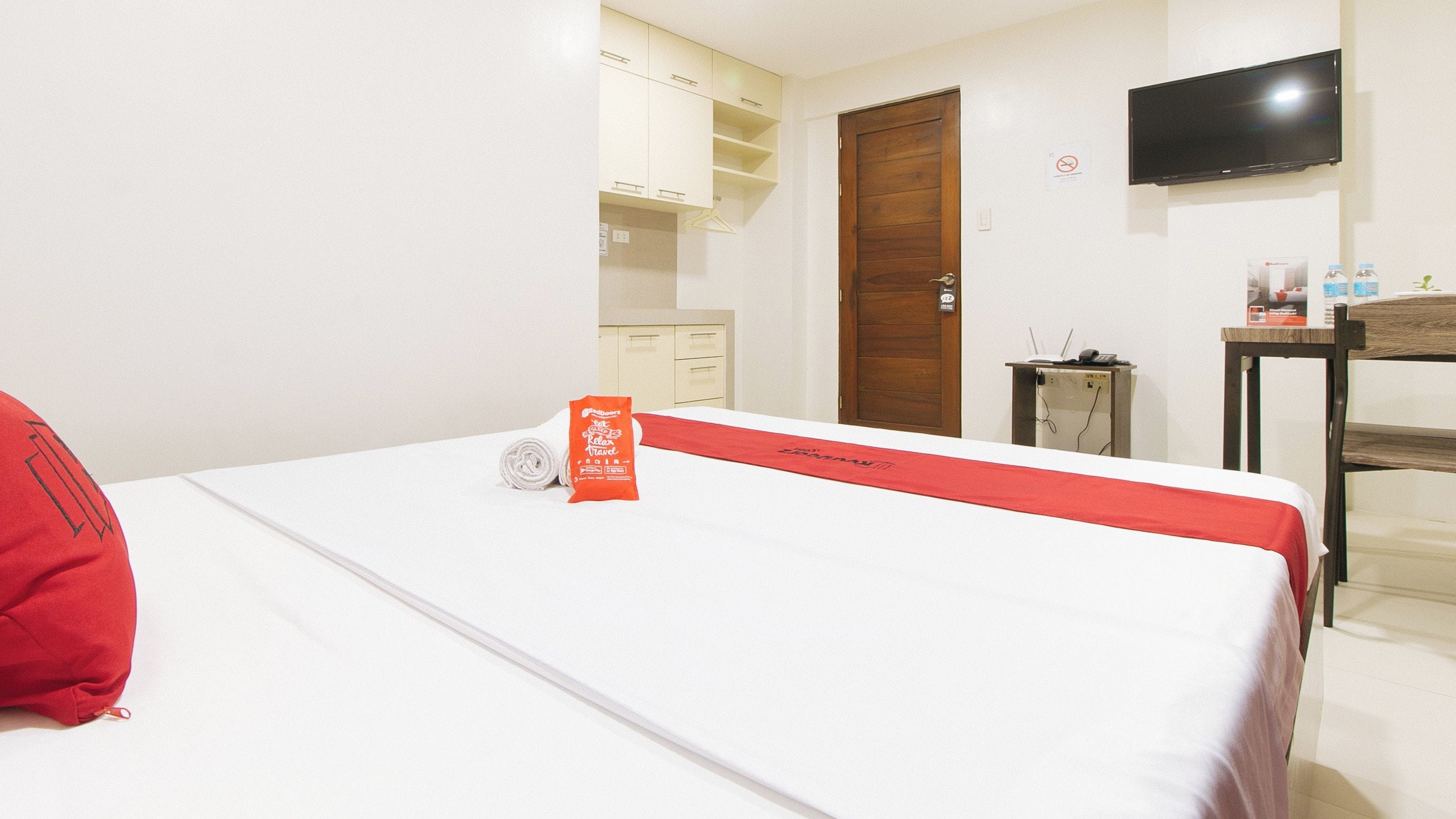 Quarto de hotel com cama, porta de madeira, TV fixada na parede e uma mesinha com uma garrafa e uma caixa.