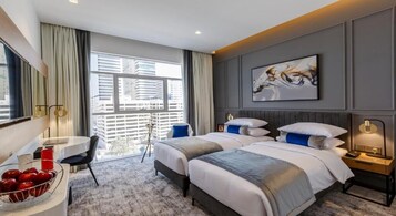 Quarto de hotel com duas camas, uma escrivaninha, uma cadeira, janela com cortinas e um quadro na parede.